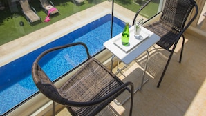 Uma varanda com piscina, duas cadeiras de vime, uma mesa pequena com garrafa e copo, e vista para um jardim e área de piscina.