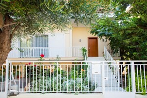 Um edifício residencial com varanda, escadaria e área de jardim com diversas plantas e uma rosa vermelha.