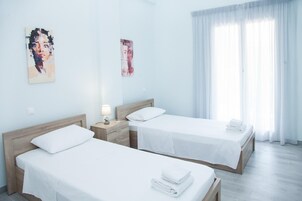 Quarto de hotel com duas camas de solteiro, uma mesa de cabeceira com abajur e uma janela grande com cortinas translúcidas.