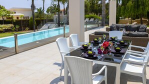 Uma área de refeições à beira da piscina com uma mesa posta para dois, rodeada por cadeiras de vime branco, com vista para uma piscina e palmeiras.