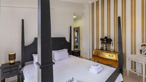 Uma cama com dossel com lençóis brancos e um edredom acolchoado, uma mesinha de cabeceira de madeira com abajur, um penteou dourado com espelho e uma cadeira branca com almofada.