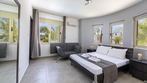 Quarto de hotel moderno com uma cama grande, um sofá, uma mesinha de cabeceira e uma janela com vista para palmeiras.