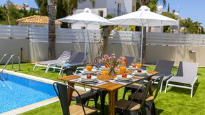 Uma área de refeições à beira da piscina, com uma mesa posta para dois, duas guarda-sóis e a piscina.