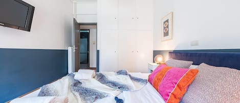 Quarto de hotel com uma cama, mesa de cabeceira com abajur, televisão na parede e porta que dá para outro quarto.