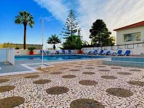 Uma área de piscina com piso de azulejos, espreguiçadeiras e palmeiras.