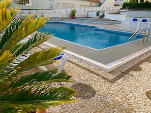 Uma piscina com deck ladrilhado, escada de metal e uma palmeira em primeiro plano.