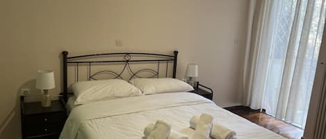 Cama bem arrumada com lençóis brancos e toalhas dobradas, cabeceira em estrutura de metal, abajures laterais e janela com cortinas leves.