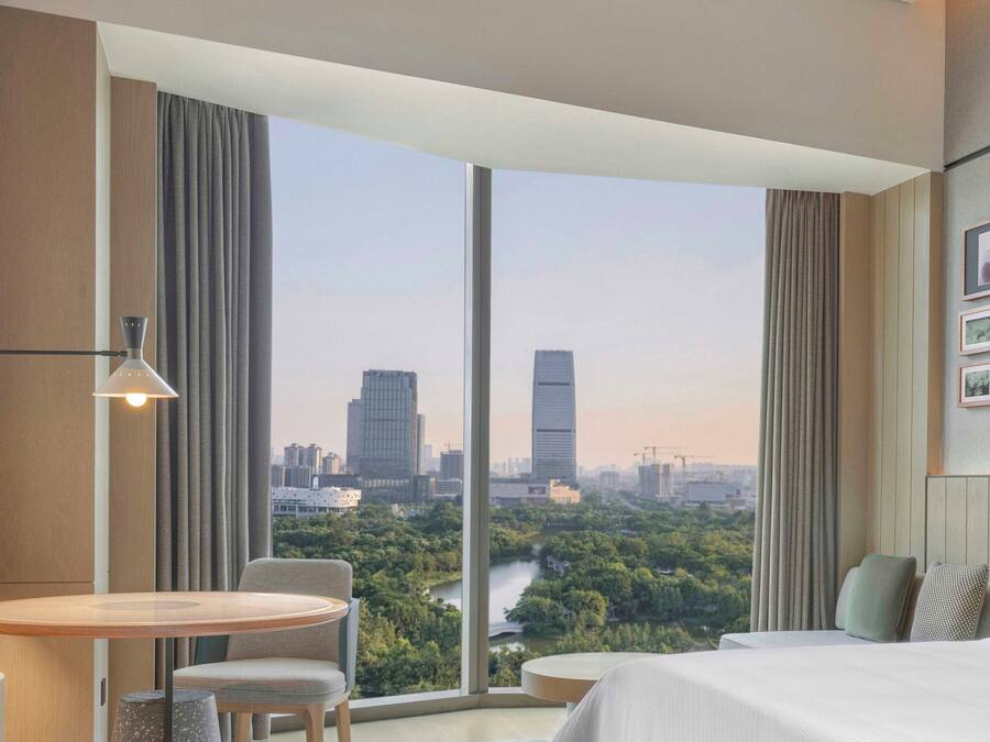 Quarto de hotel moderno com uma grande janela com vista para a cidade, uma mesa redonda de madeira, uma cadeira confortável, uma cama e uma luminária fixada na parede.