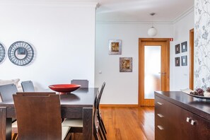 Uma sala de jantar com mesa de madeira, cadeiras e uma tigela vermelha. Há decorações nas paredes e uma porta que dá para outro cômodo.