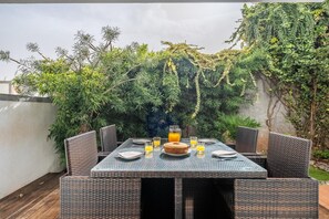 Uma área de refeições ao ar livre com móveis de vime e uma mesa de vidro, cercada por vegetação exuberante.
