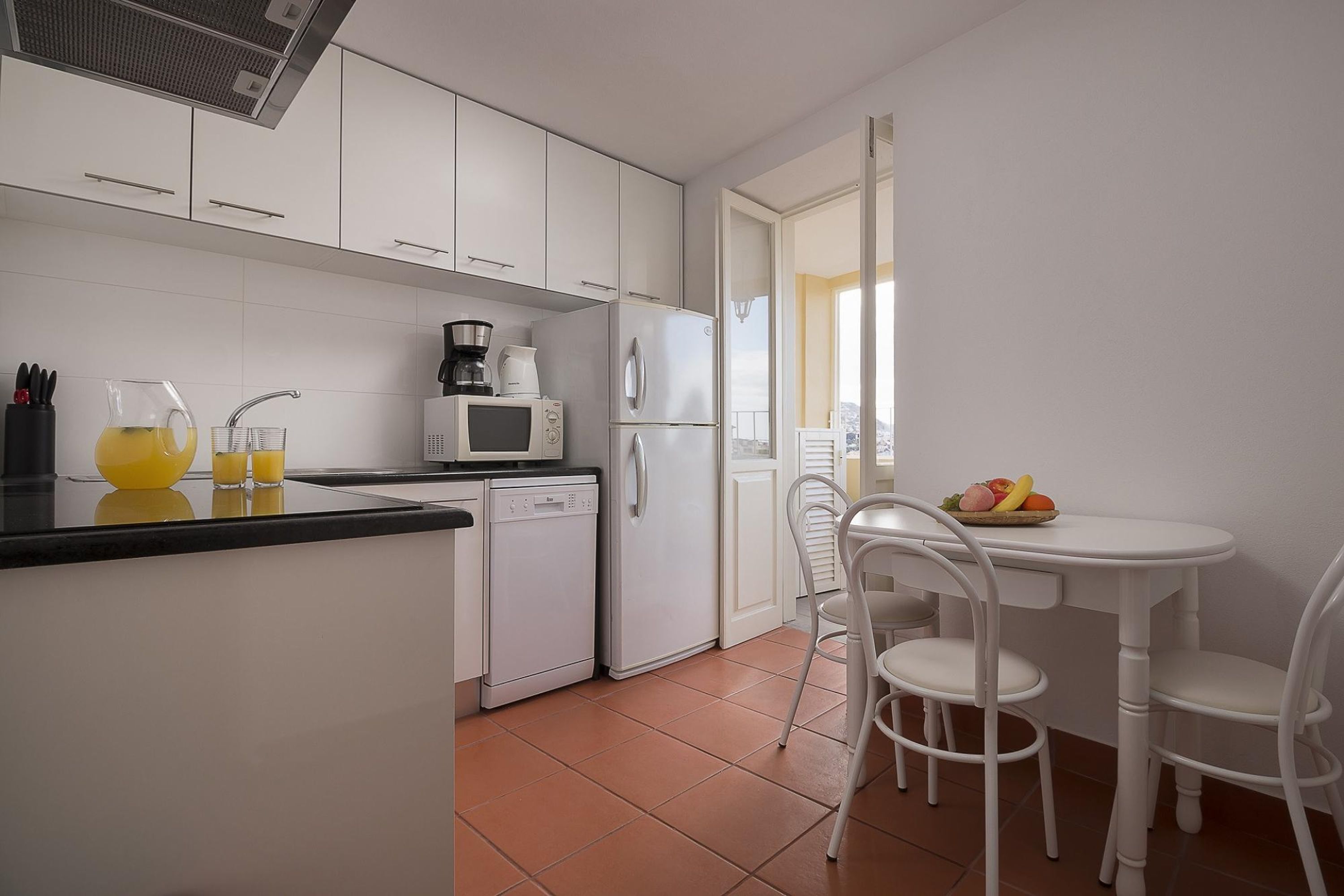 Cozinha compacta com armários brancos, micro-ondas, geladeira, cafeteira e uma pequena mesa de jantar com cadeiras.