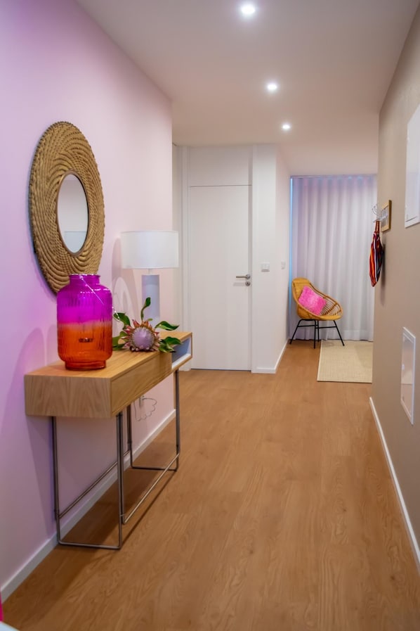 Um corredor com chão de madeira, um espelho, uma mesa com um vaso, um abajur e uma porta que dá para outro quarto.