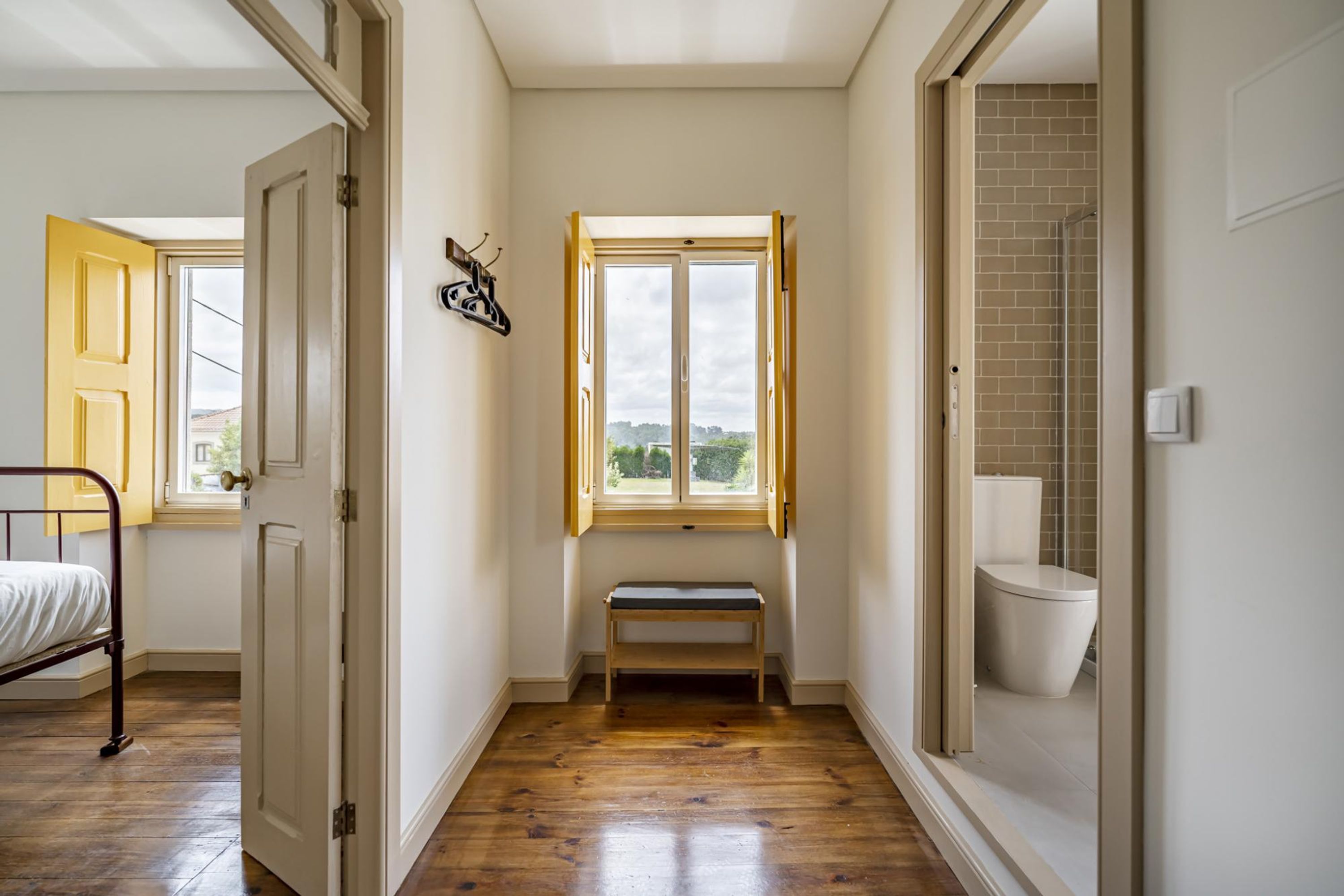 Um corredor estreito com piso de madeira, um banco, uma janela com vista e uma porta para um banheiro.