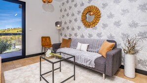 Sala de estar com sofá, mesa de centro, abajur, espelho decorativo e vista através de uma porta de vidro.