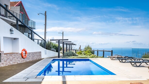 Piscina no terraço com vista clara do oceano, cercada por um prédio branco com telhado vermelho e um deck de madeira com espreguiçadeiras.