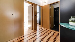 Um corredor com piso de madeira e padrão quadriculado, que leva a um quarto com uma porta preta e um objeto branco sobre um balcão verde-água.