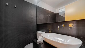 Um banheiro moderno com uma pia grande e branca, paredes revestidas de azulejos escuros, vaso sanitário e espelho com moldura dourada.