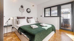 Quarto com cama, mesas de cabeceira, janela com persianas e vista para o exterior.