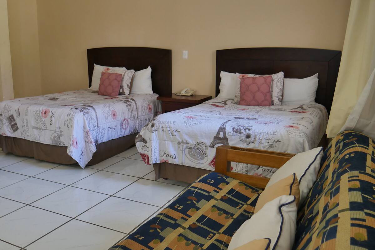 Duas camas com cabeceiras iguais e roupa de cama decorativa, uma cadeira e um sofá estampado em um quarto com piso de azulejos.