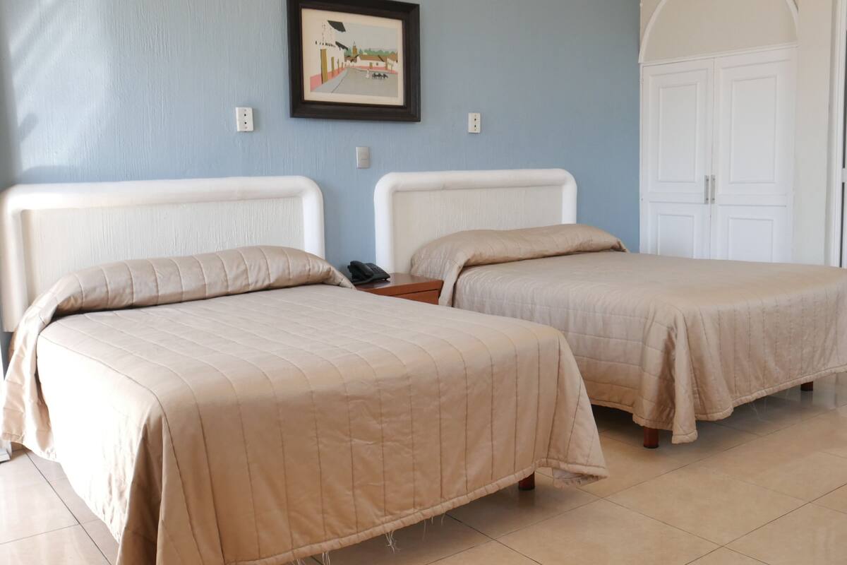 Duas camas de solteiro com roupa de cama bege, uma pequena mesa de cabeceira com telefone, um quadro emoldurado na parede e uma porta branca ao fundo.