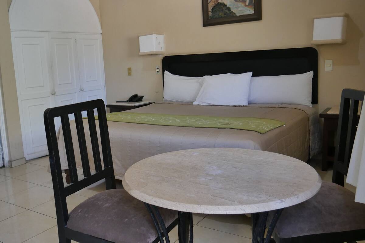 Quarto de hotel com cama, mesa redonda, duas cadeiras, uma porta e um quadro na parede.