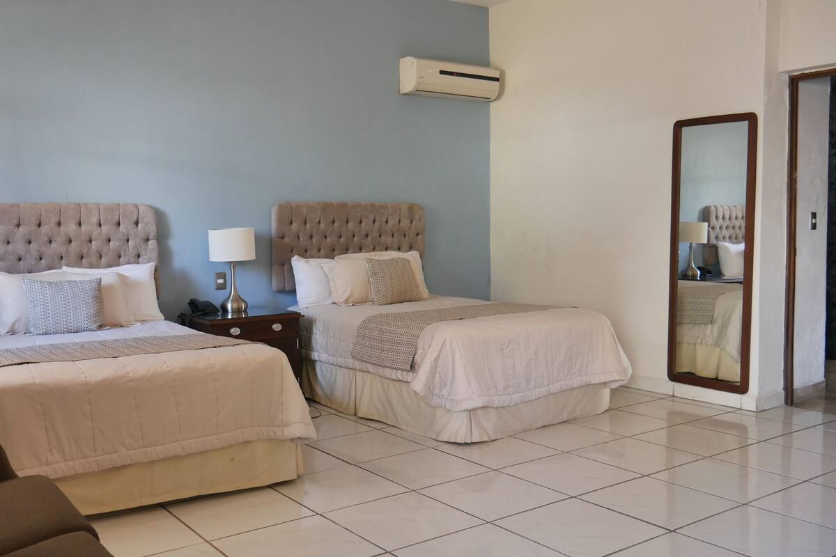 Quarto de hotel com duas camas, uma mesa de cabeceira, um abajur, aparelho de ar condicionado e um espelho.