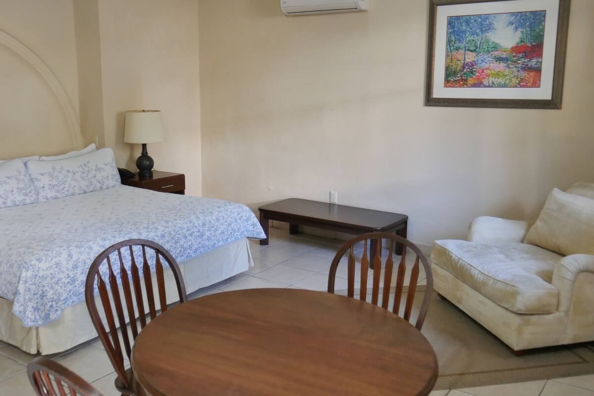 Quarto com cama, mesa de cabeceira, abajur, cadeira, sofá e uma pequena mesa.