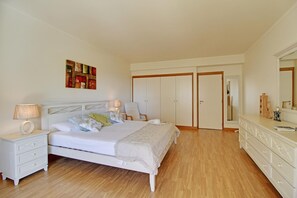 Quarto com cama, mesas de cabeceira, cômoda, espelho e porta que dá para outro espaço.