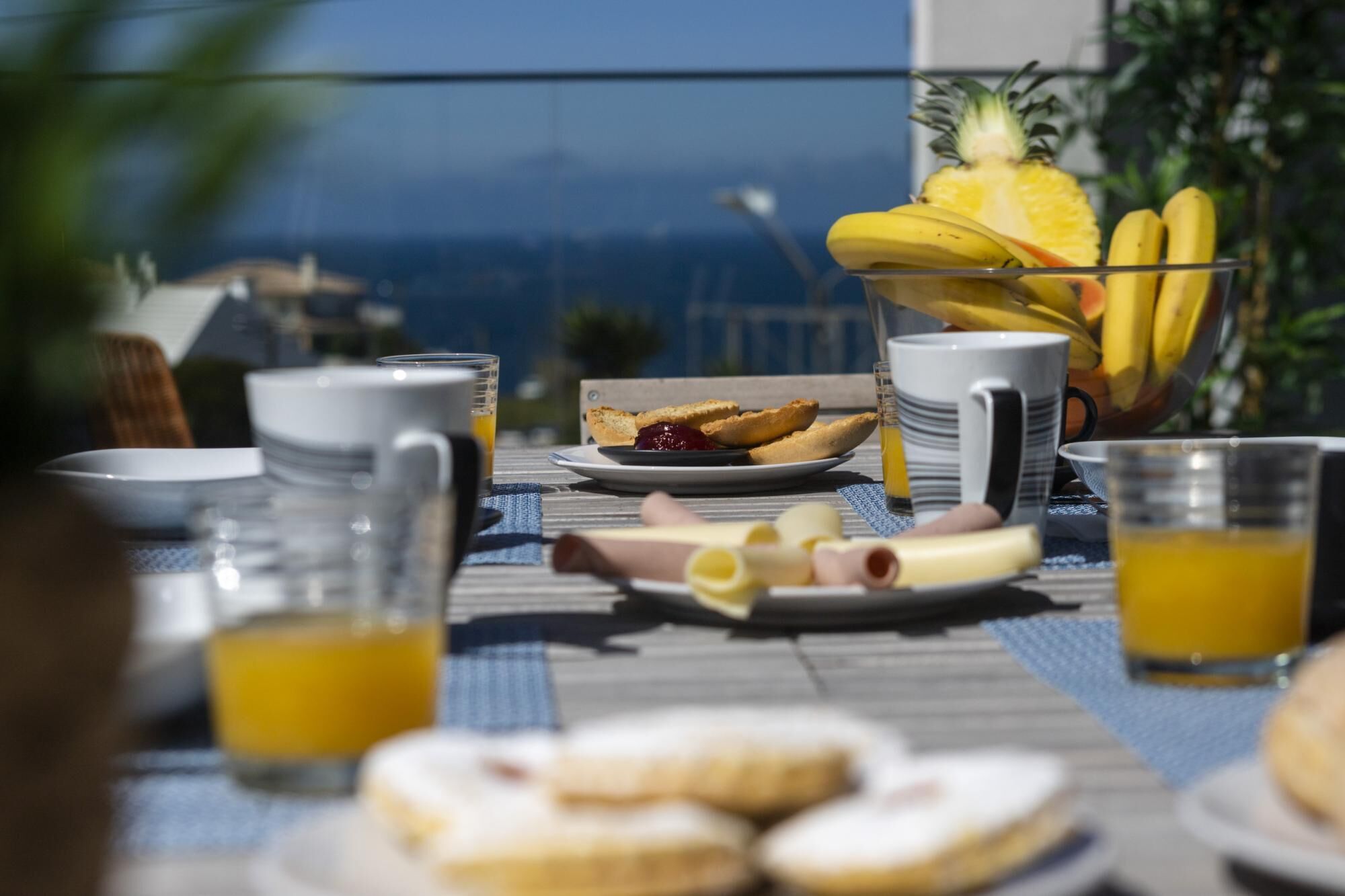 Uma mesa de café da manhã com diversos alimentos, incluindo pães, doces e frutas, além de copos e taças de suco de laranja.