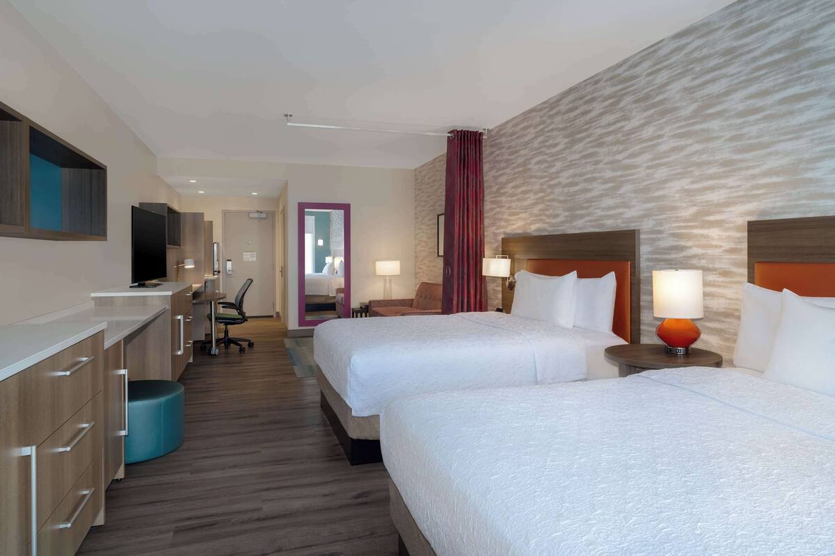 Quarto de hotel com duas camas, uma escrivaninha, uma televisão e um banheiro visível por uma porta aberta.