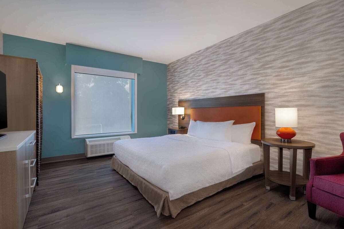 Quarto de hotel com uma cama grande, uma escrivaninha, uma cadeira e uma janela com persianas.
