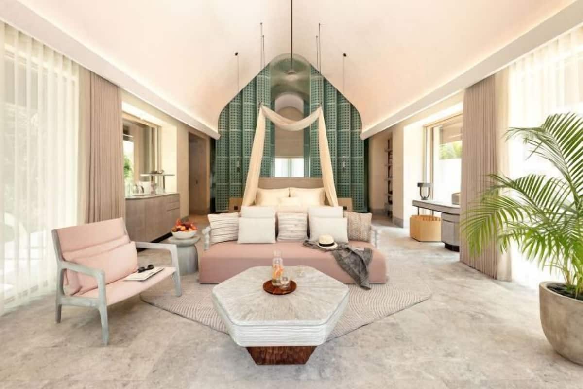 Quarto de hotel moderno com cama central, área de estar com sofá e poltronas, mesa de centro e janelas amplas.