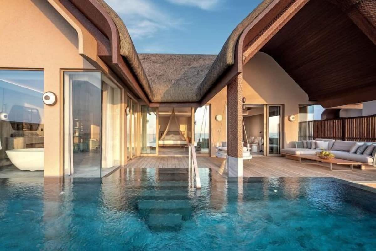 Uma piscina moderna de borda infinita com barreira de vidro, deck de madeira e área de descanso com sofá.