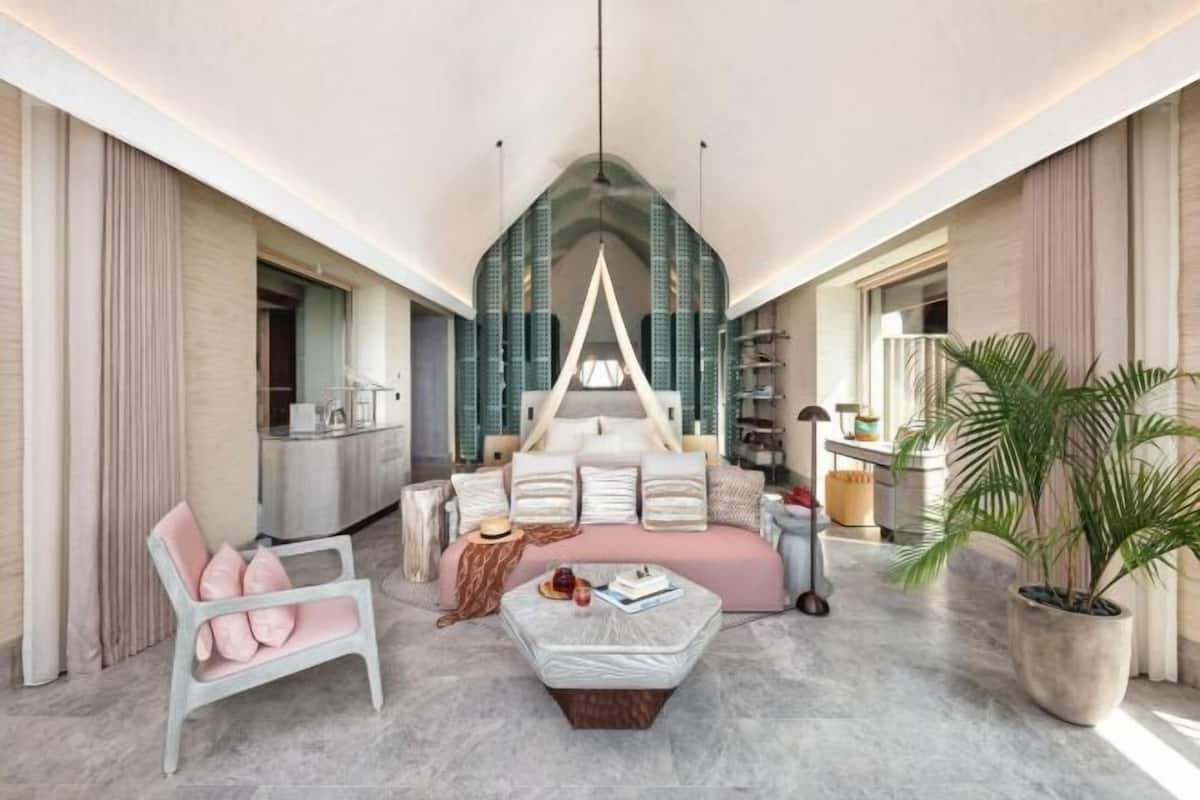 Quarto de hotel moderno com cama central, área de estar com uma cadeira e uma mesa, e uma grande planta em vaso.