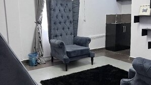 Sala de estar com poltrona cinza com capitonê, tapete preto e área de cozinha com armários.