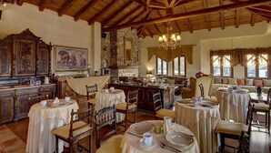 Uma sala de jantar com vigas de madeira, parede de pedra e um candelabro. Mesas postas com toalhas brancas e talheres.