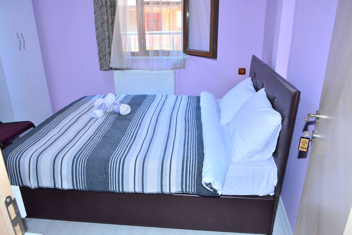 Uma cama bem arrumada com roupa de cama listrada e almofadas brancas, uma janela com cortinas finas e uma porta com placa de número do quarto.