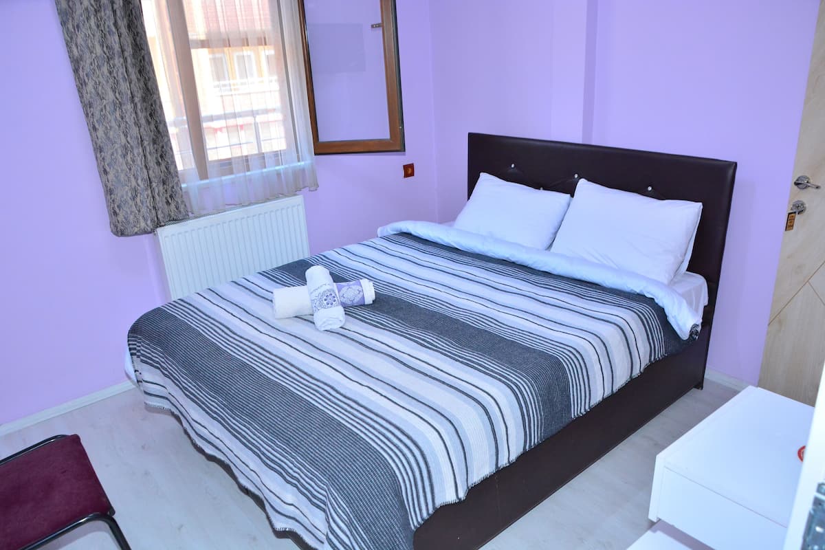 Uma cama bem arrumada com roupa de cama listrada, uma mesa de cabeceira branca e uma janela com cortinas leves.