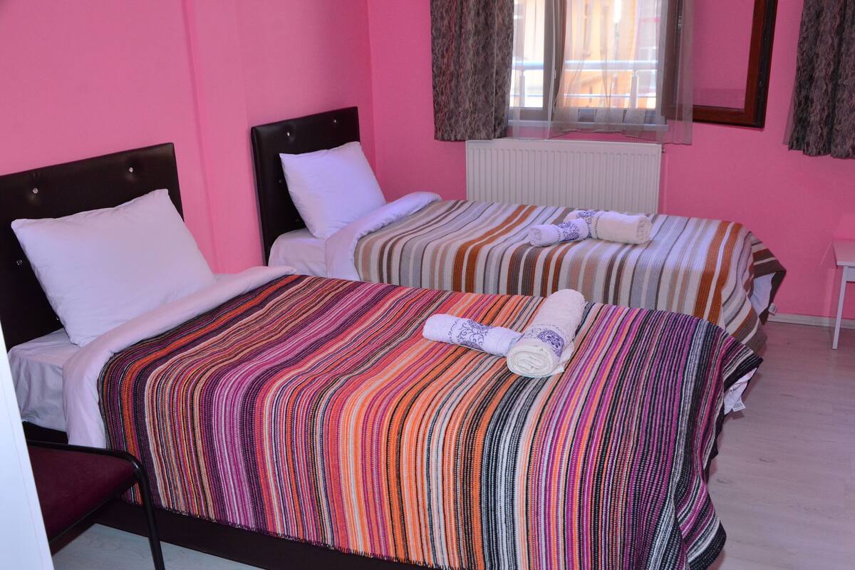 Quarto de hotel com duas camas, cada uma com colcha listrada e almofadas brancas. Há uma janela com cortinas, um radiador e uma pequena mesa ao lado das camas.