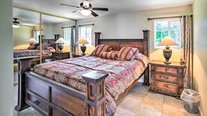 Quarto com uma cama grande, um ventilador de teto, mesa de cabeceira com abajur e janela com cortinas.