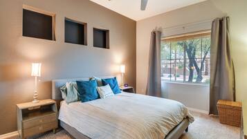 Quarto com cama, mesas de cabeceira, ventoinha no teto, janela com cortinas e vista para árvores.