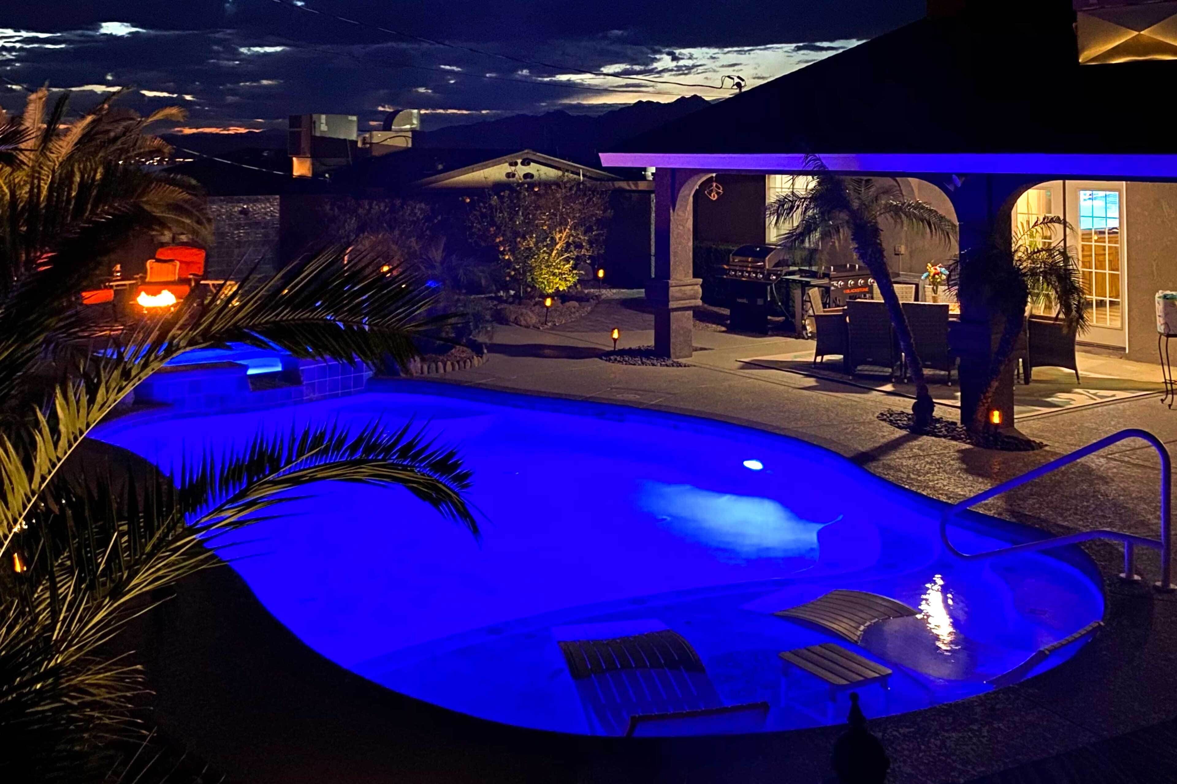 Piscina iluminada em azul, rodeada por palmeiras e um espaço de terraço com mobiliário de exterior.