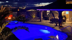 Piscina iluminada em azul, rodeada por palmeiras e um espaço de terraço com mobiliário de exterior.