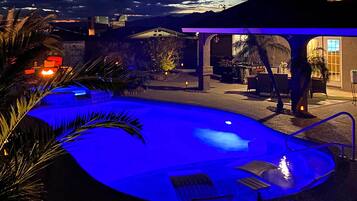 Piscina iluminada em azul, rodeada por palmeiras e um espaço de terraço com mobiliário de exterior.