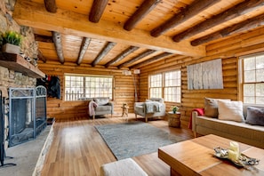 Interior de cabana de toras com lareira, piso de madeira, sofá e mesa de centro.