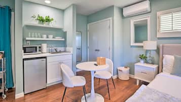 Quarto de hotel compacto com cozinha compacta, uma mesa de jantar redonda com duas cadeiras, uma cama com edredom branco, uma mesa de cabeceira com abajur e um espelho grande na parede.