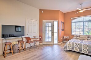 Quarto com cama, televisão, área de jantar com cadeiras e mesa, e porta que dá para o exterior.