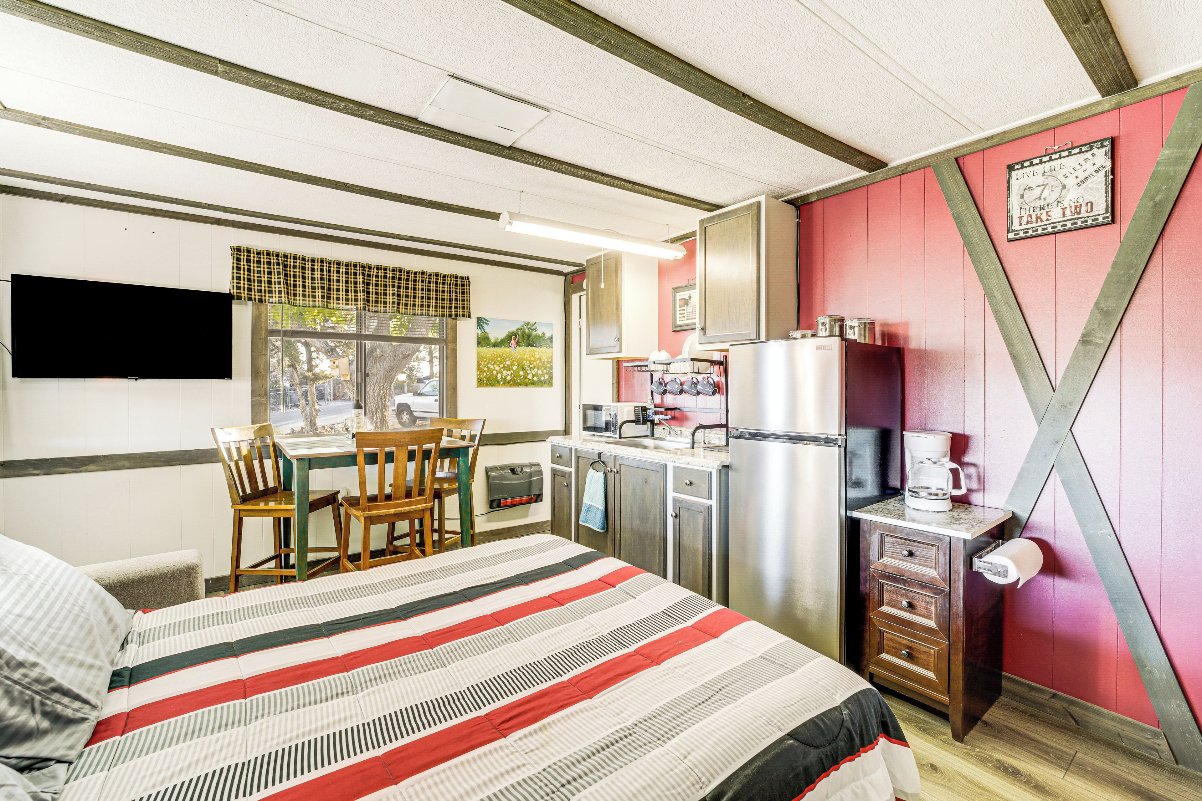 Quarto compacto com cama, pequena mesa de jantar com cadeiras, cozinha compacta com geladeira e televisão fixada na parede.