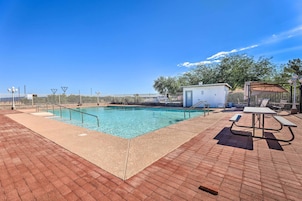 Piscina retangular com guarda-corpo de metal, rodeada por um terraço de tijolos. Há espreguiçadeiras e um pequeno edifício com porta, provavelmente um vestiário ou depósito.