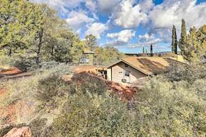 Uma casa rodeada por vegetação densa e árvores, com céu claro e uma pequena construção nas proximidades.